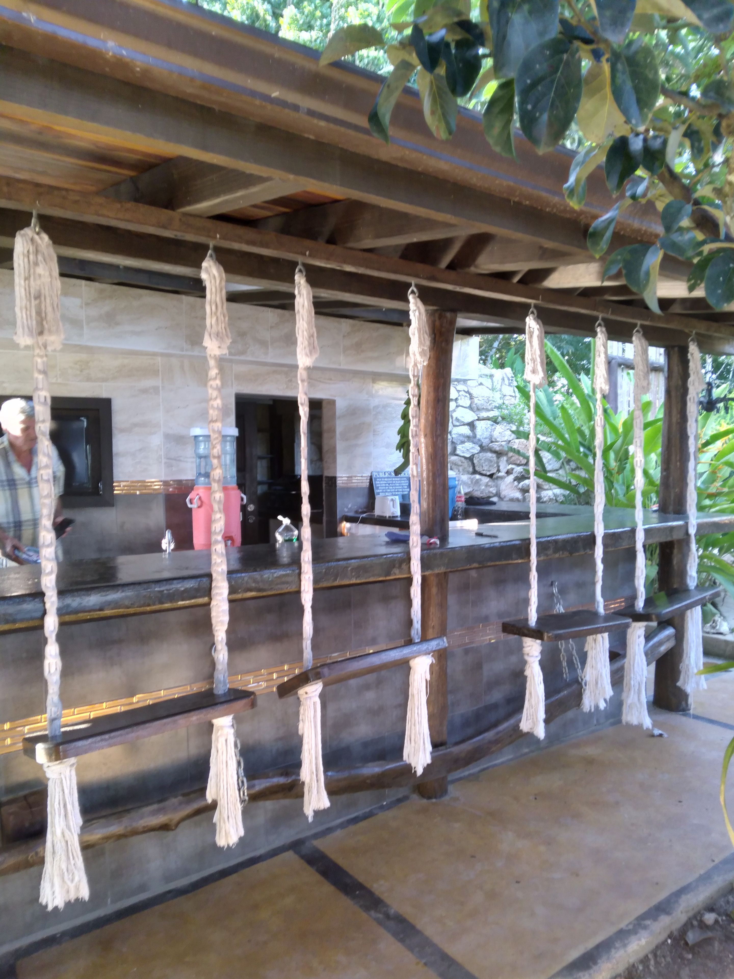 Área externa coberta com parede de pedra, balcão com objeto vermelho e detalhes decorativos em macramé.