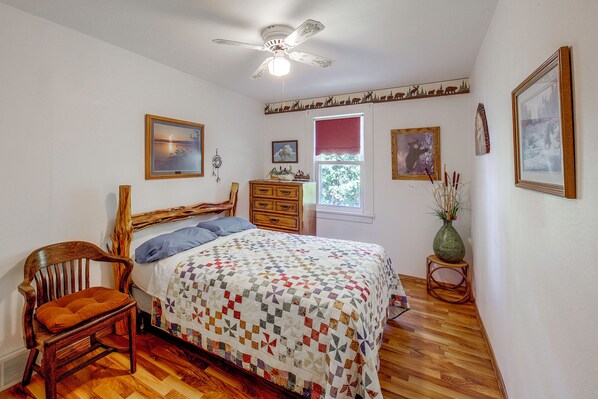 Quarto com cama de madeira, uma cadeira, cômoda e ventilador de teto.