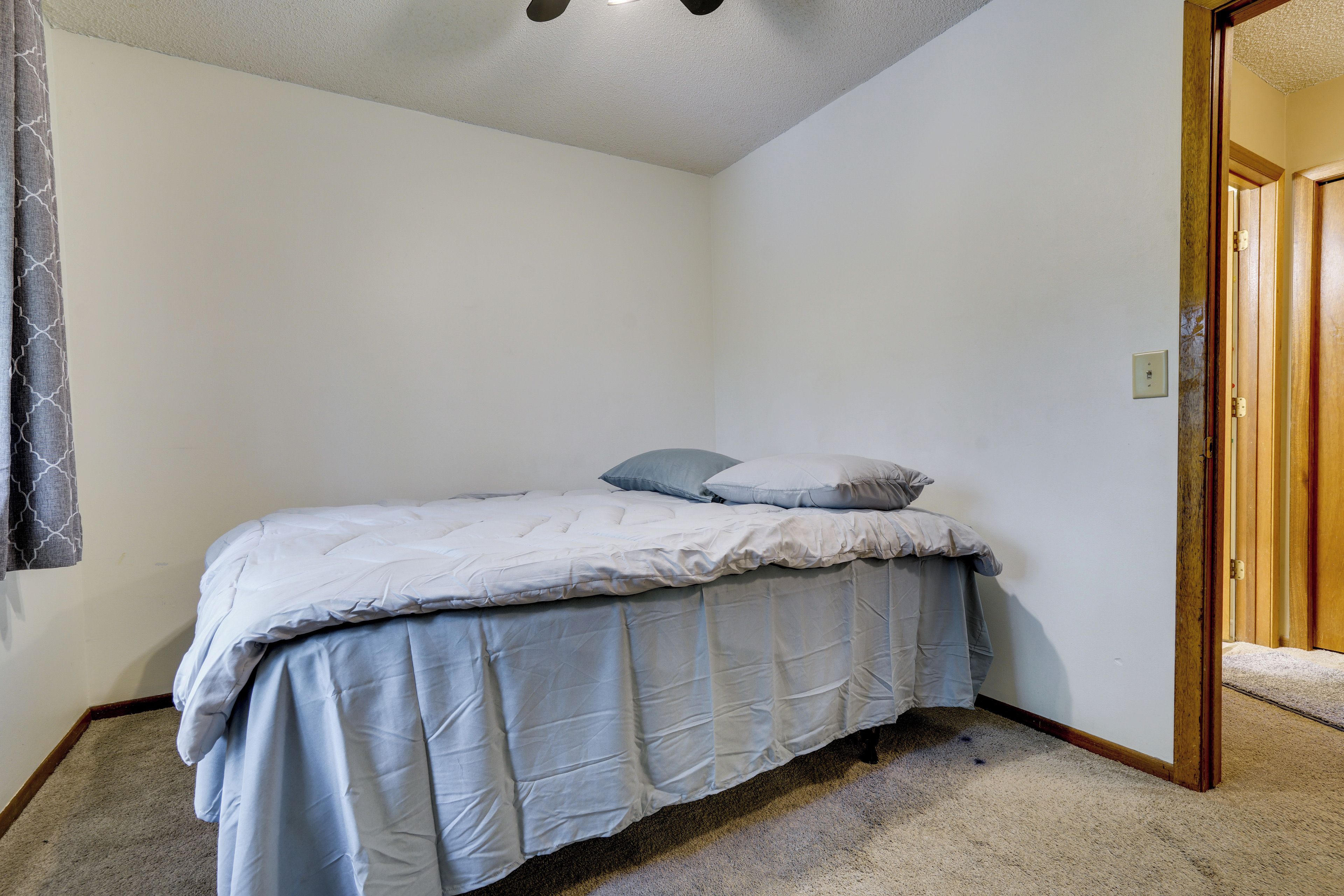 Quarto pequeno com uma cama de solteiro, parede branca lisa, porta de madeira e chão coberto de carpete.