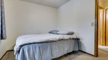 Quarto pequeno com uma cama de solteiro, parede branca lisa, porta de madeira e chão coberto de carpete.