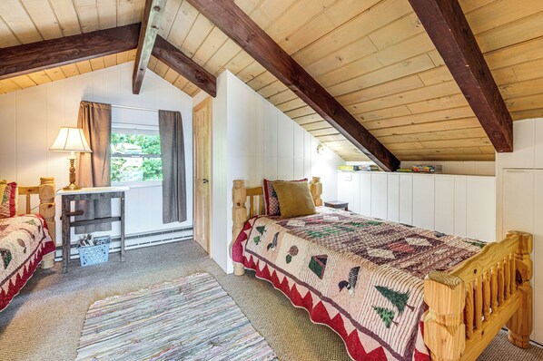 Quarto aconchegante com teto inclinado, cama com colcha estampada, mesa de cabeceira com abajur e janela com cortinas.