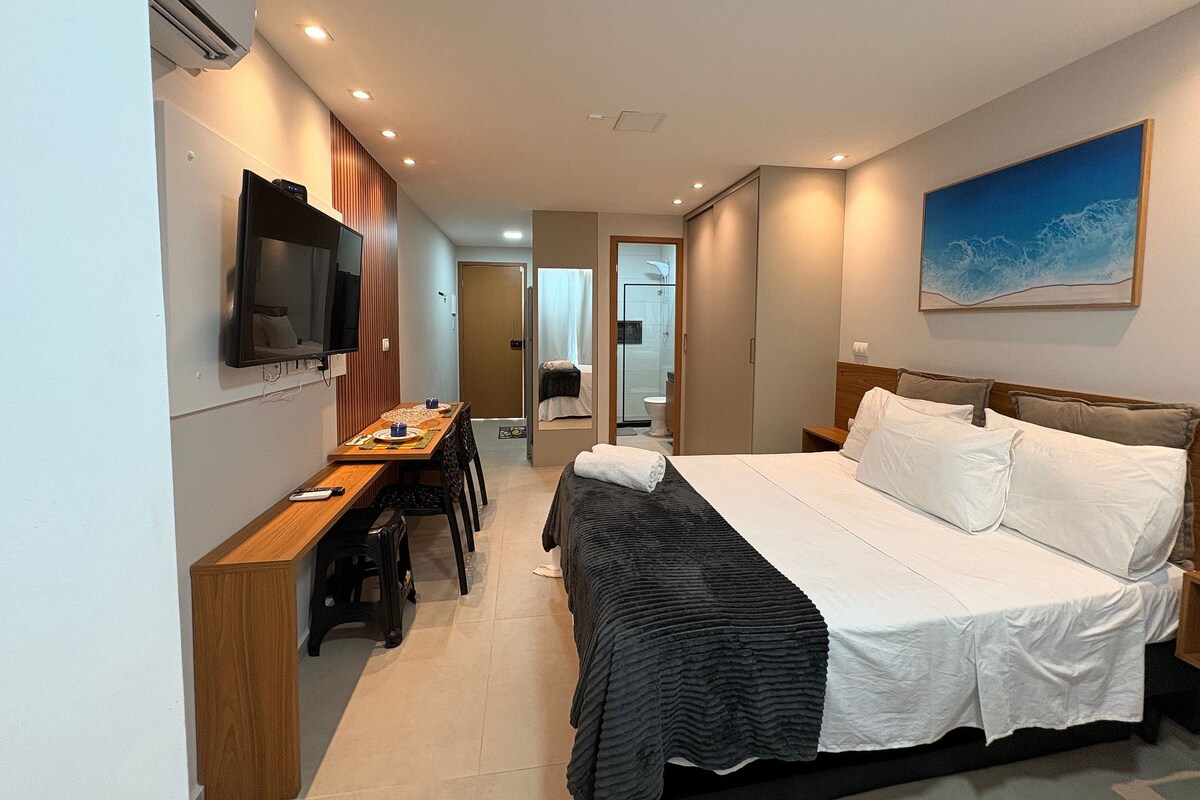 Quarto de hotel com cama, escrivaninha, cadeira, televisão e um quadro na parede.