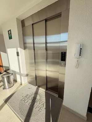 Elevador moderno com acabamento metálico, parede texturizada e uma pequena lixeira à esquerda.