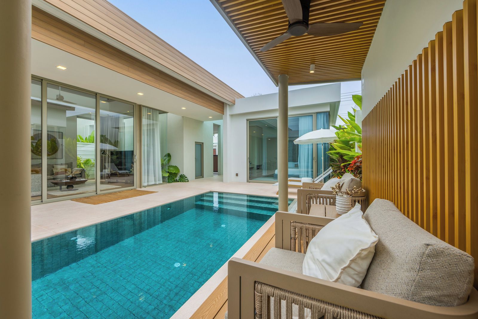 Uma área de piscina moderna com uma sala envidraçada, deck de madeira e um espaço de descanso com sofá e almofadas.