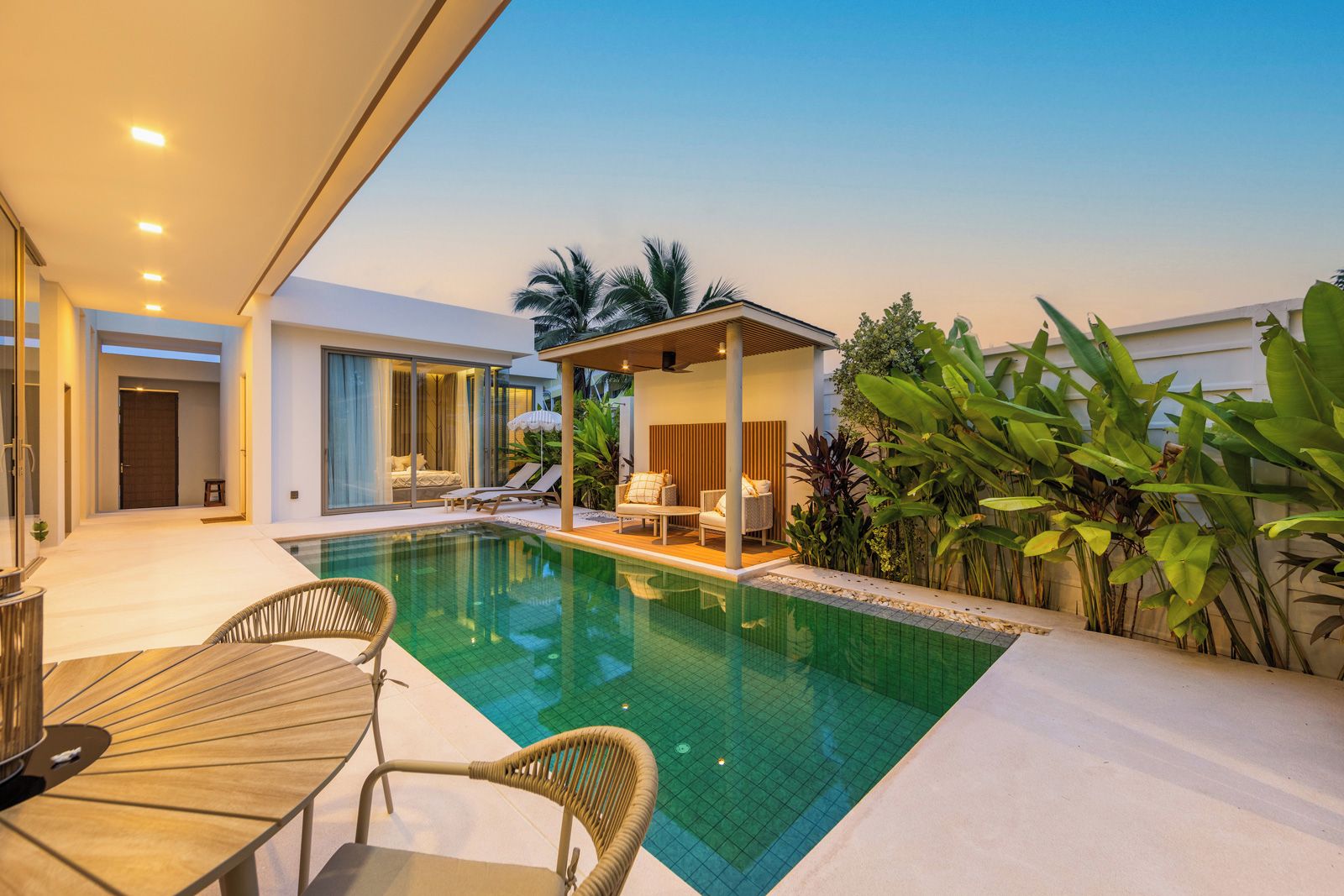 Uma área de piscina moderna com espreguiçadeiras e um espaço coberto para sentar, rodeada por plantas tropicais.