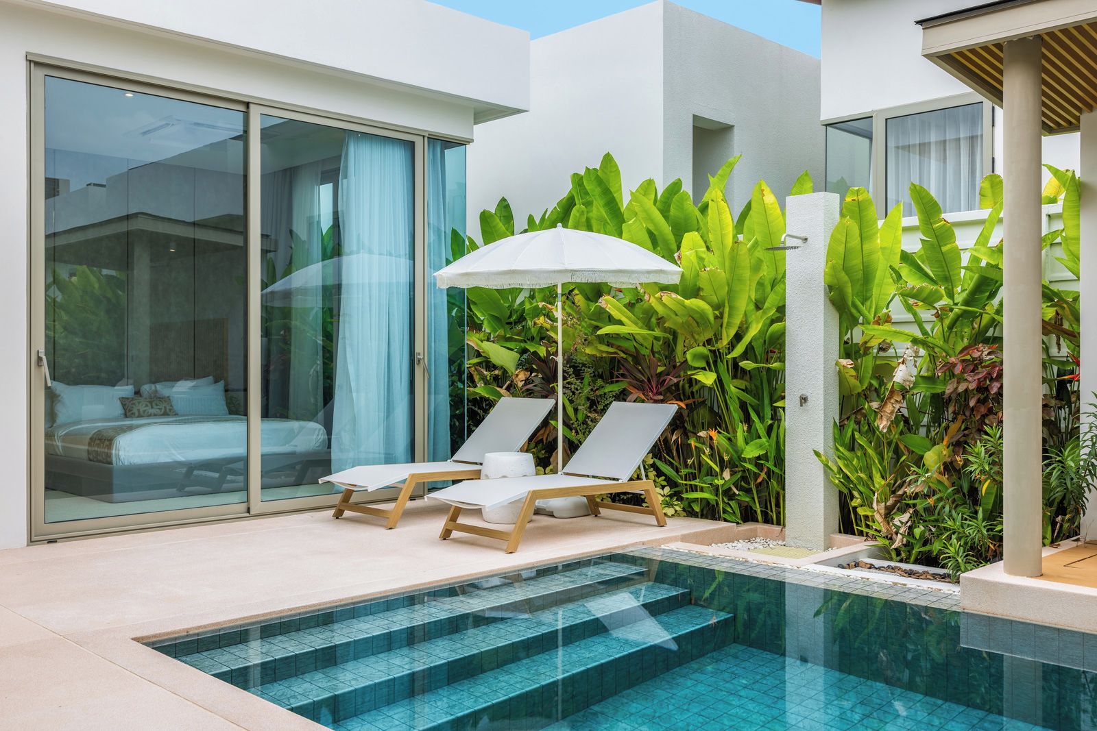 Área de piscina moderna ao ar livre, com duas espreguiçadeiras, um quarto com paredes de vidro e plantas tropicais exuberantes.