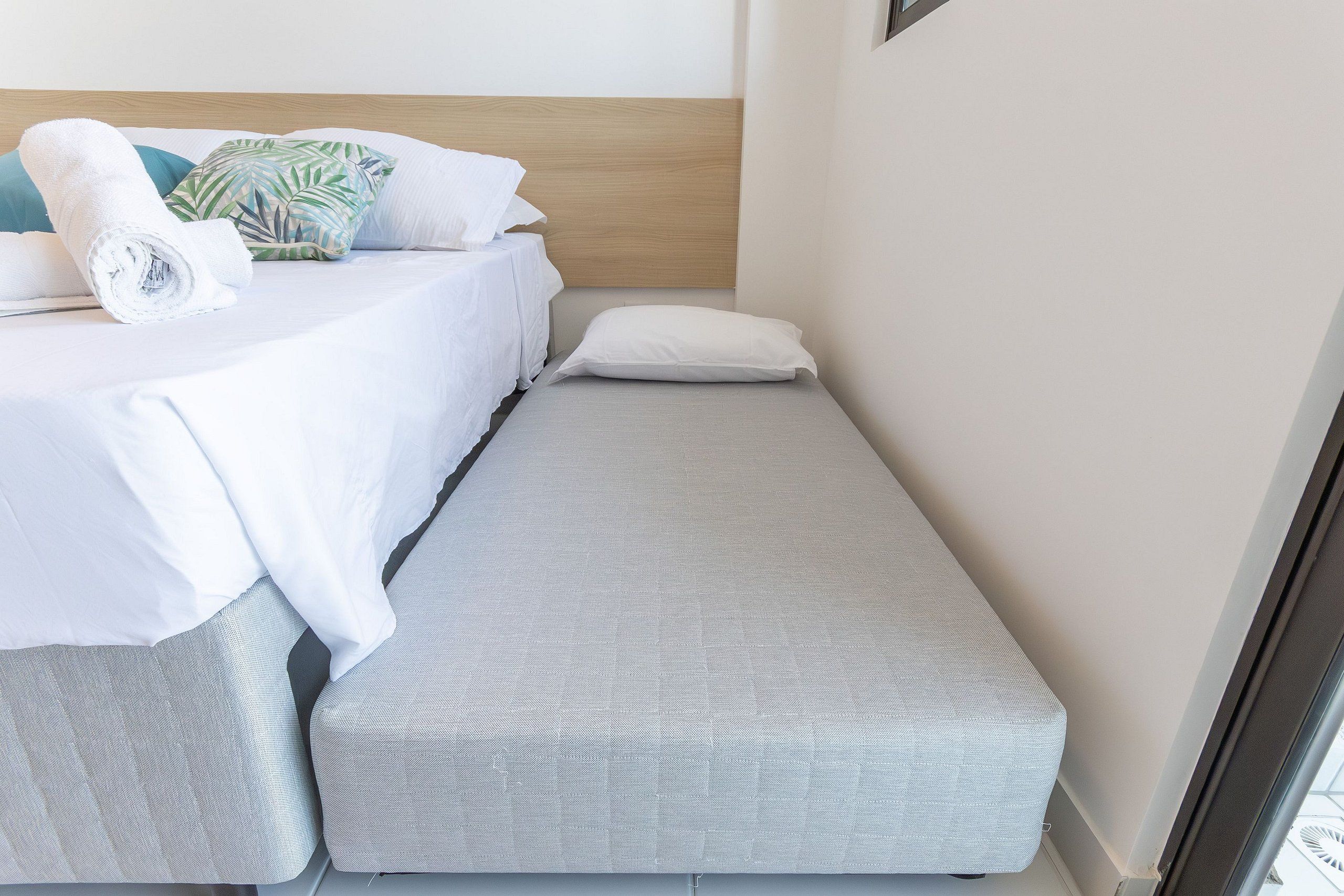 Quarto pequeno, com cama de casal, cabeceira de madeira, colcha branca e um banquinho cinza.