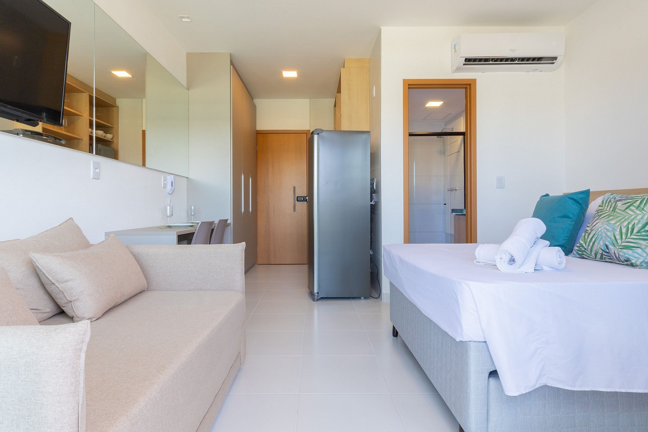 Quarto de hotel com cama, sofá, geladeira e banheiro visível por uma porta aberta.