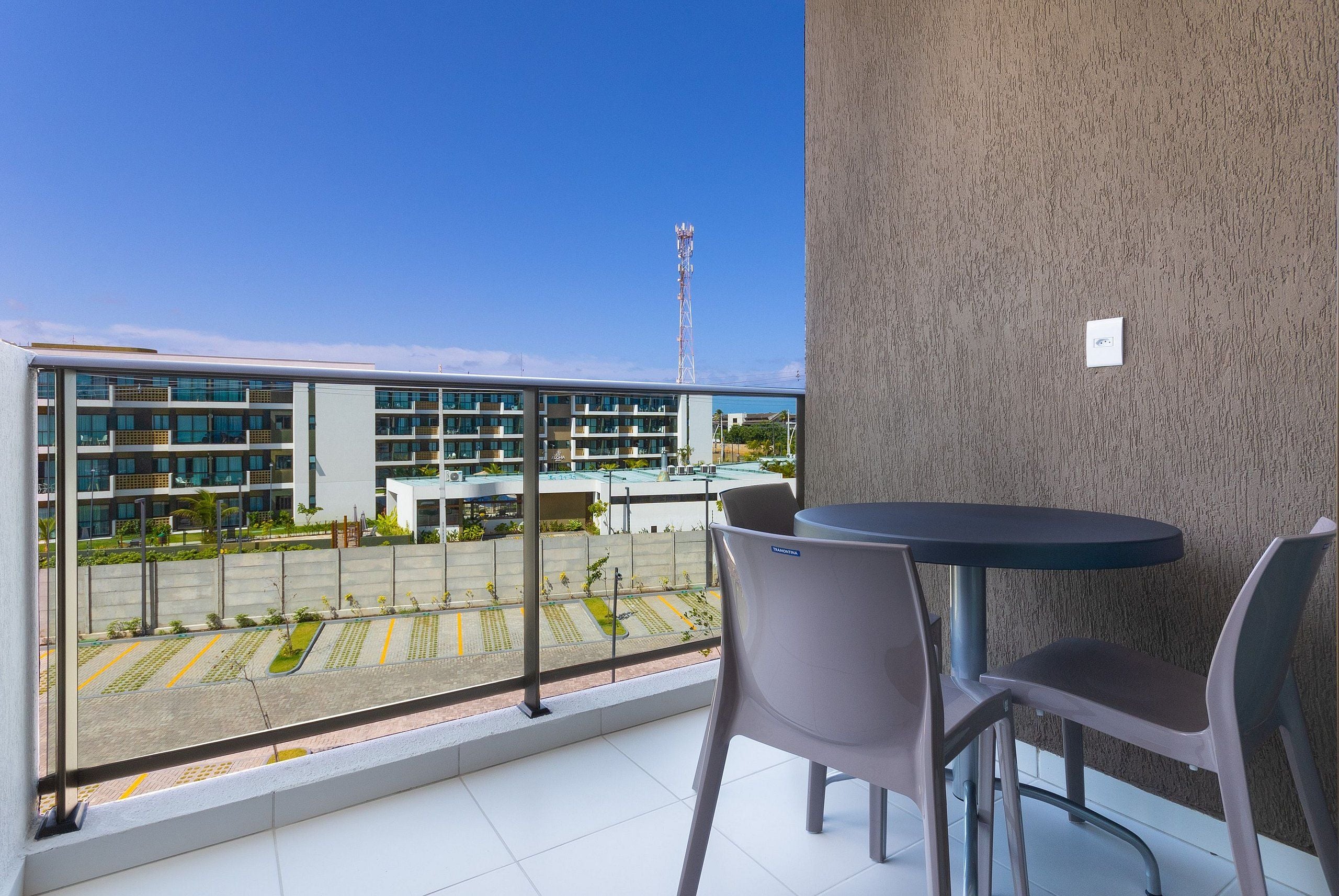 Varanda com mesa e cadeiras, com vista para um estacionamento e um edifício com vários andares.