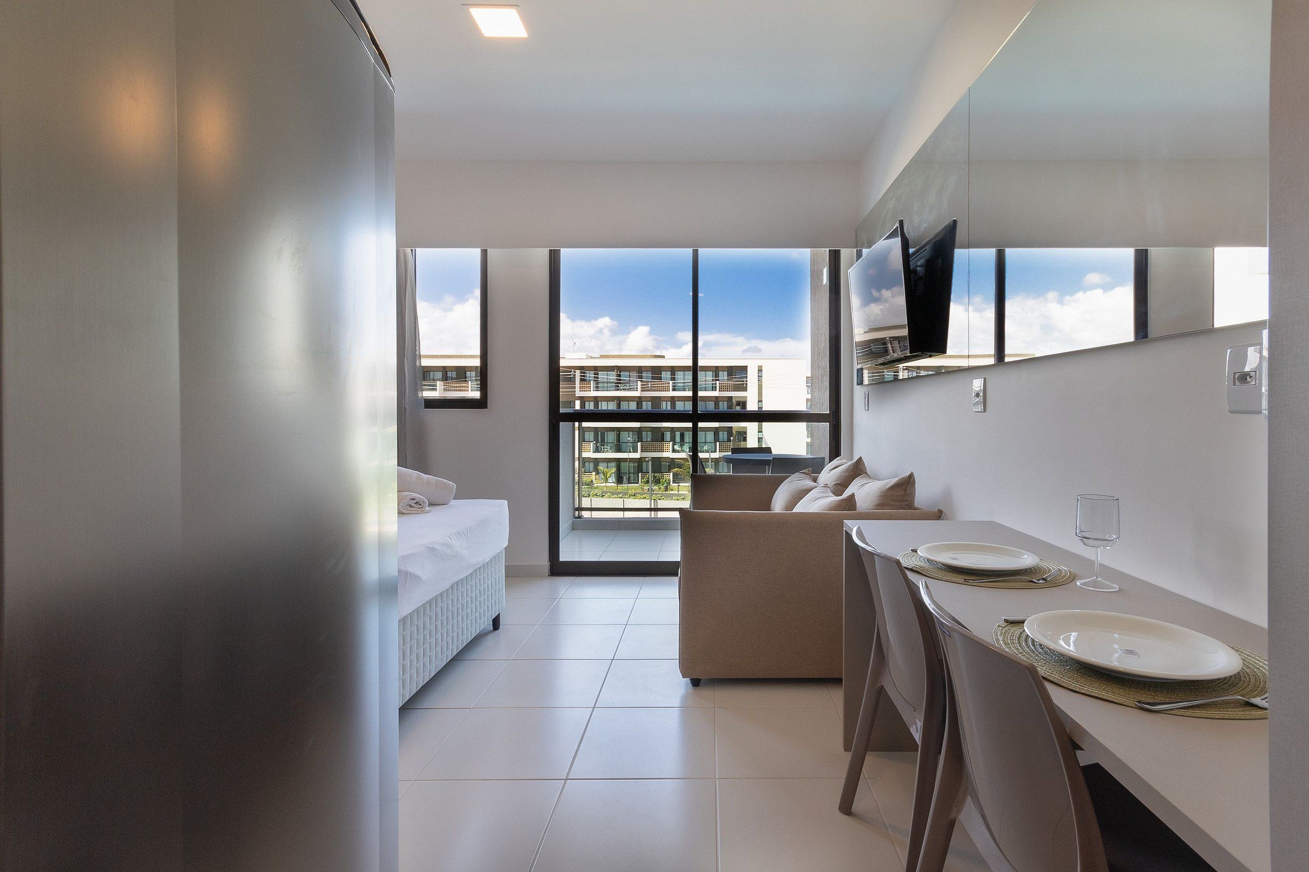 Quarto de hotel moderno com cama, sofá, área de refeições com mesa e cadeiras, e janelas amplas com vista para a cidade.