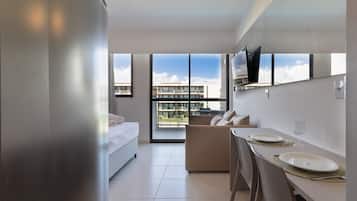 Quarto de hotel moderno com cama, sofá, área de refeições com mesa e cadeiras, e janelas amplas com vista para a cidade.