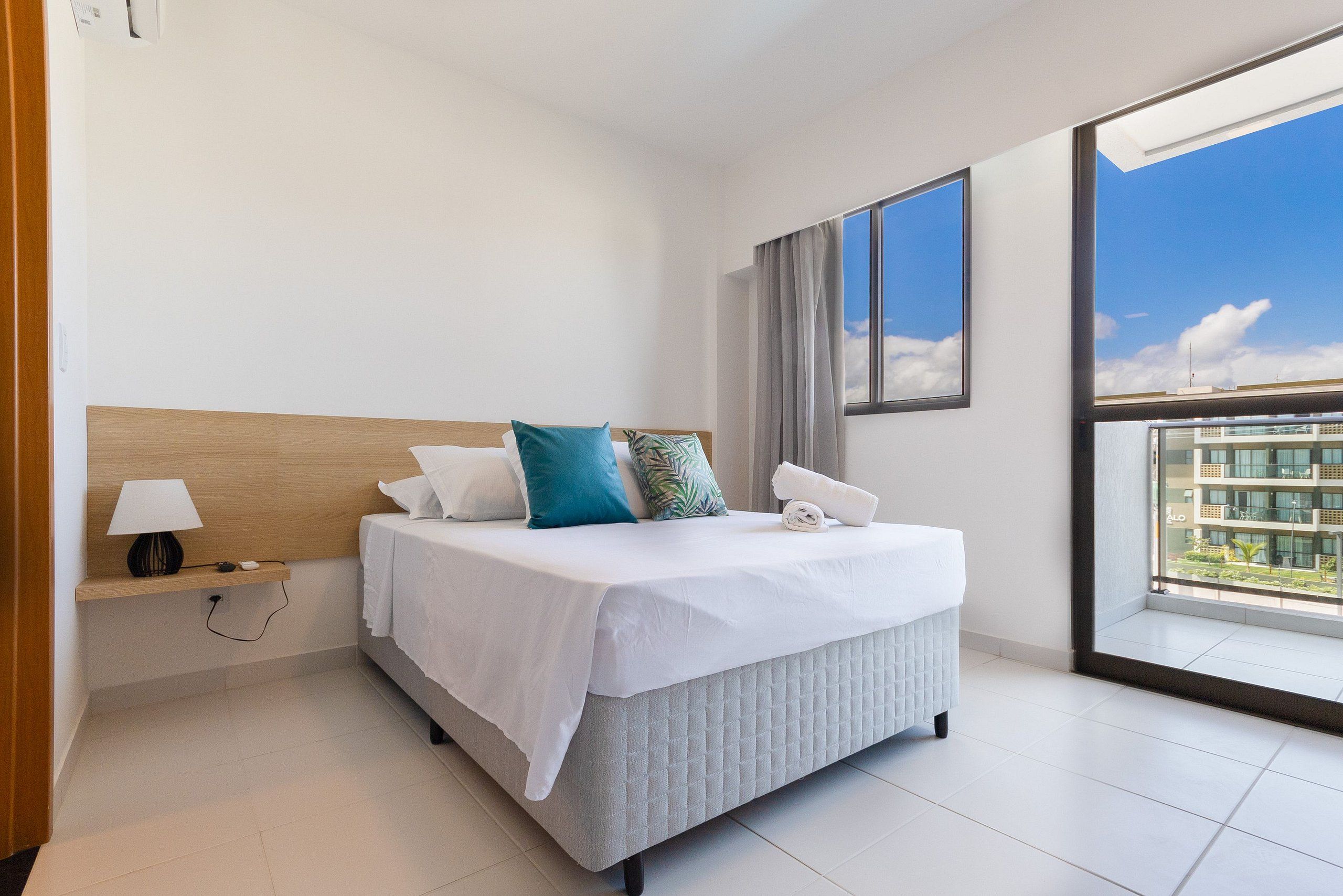 Quarto de hotel com cama, mesa de cabeceira, luminária, janela com vista para edifícios e varanda.