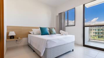 Quarto de hotel com cama, mesa de cabeceira, luminária, janela com vista para edifícios e varanda.