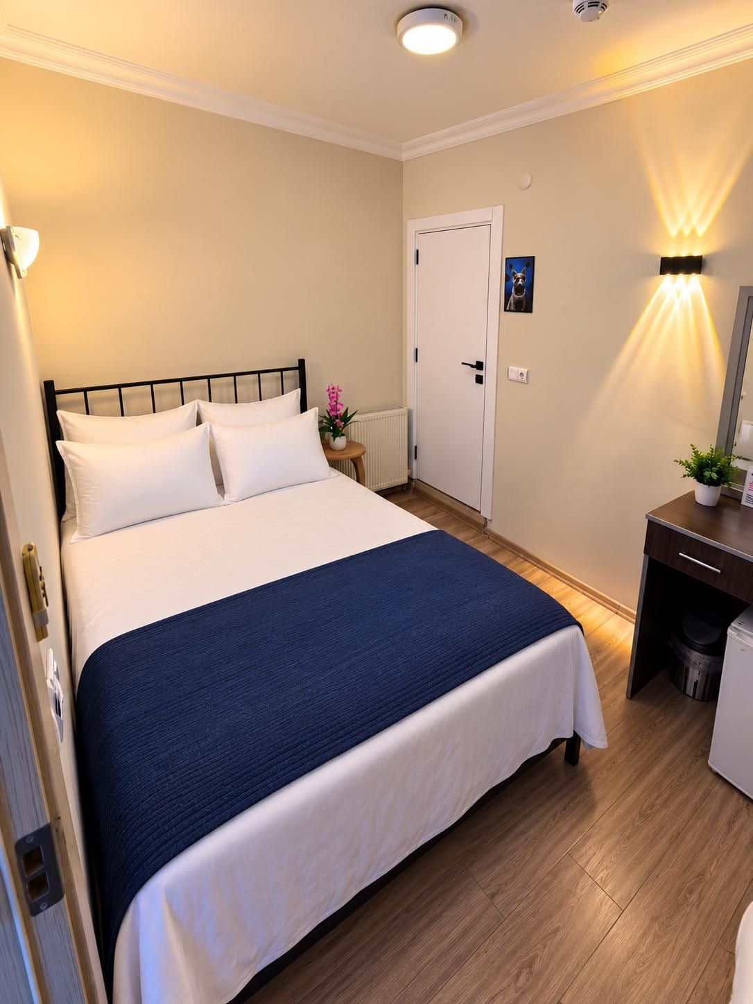 Cama bem arrumada com roupa de cama branca e azul, uma pequena mesa de cabeceira com uma planta, uma porta com um quadro de cachorro e um luminário fixado na parede.