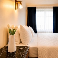 Quarto de hotel com uma cama, uma mesinha de cabeceira com um vaso de tulipas brancas e um candeeiro fixado na parede.