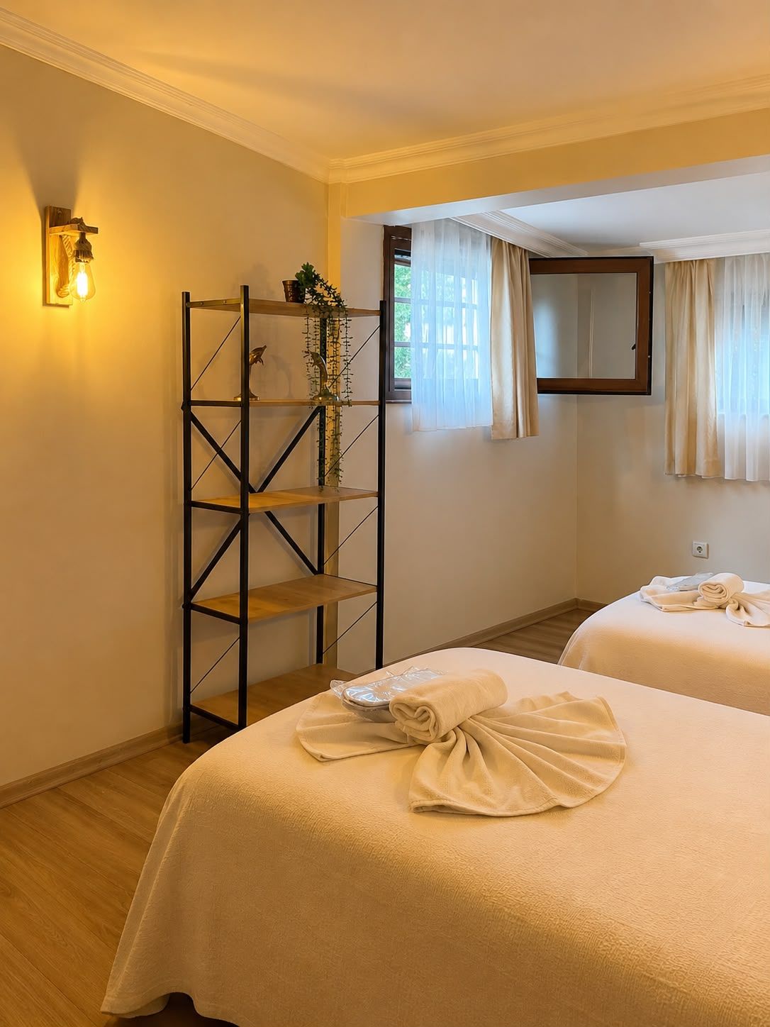 Quarto de hotel com duas camas, piso de madeira, prateleira fixa com uma planta, janela com cortinas e espelho fixado na parede.