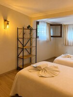 Quarto de hotel com duas camas, piso de madeira, prateleira fixa com uma planta, janela com cortinas e espelho fixado na parede.