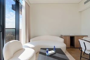 Quarto de hotel moderno com uma janela grande, um sofá, uma mesinha de centro com lenços de papel e garrafas de água, e uma escrivaninha com uma cadeira.
