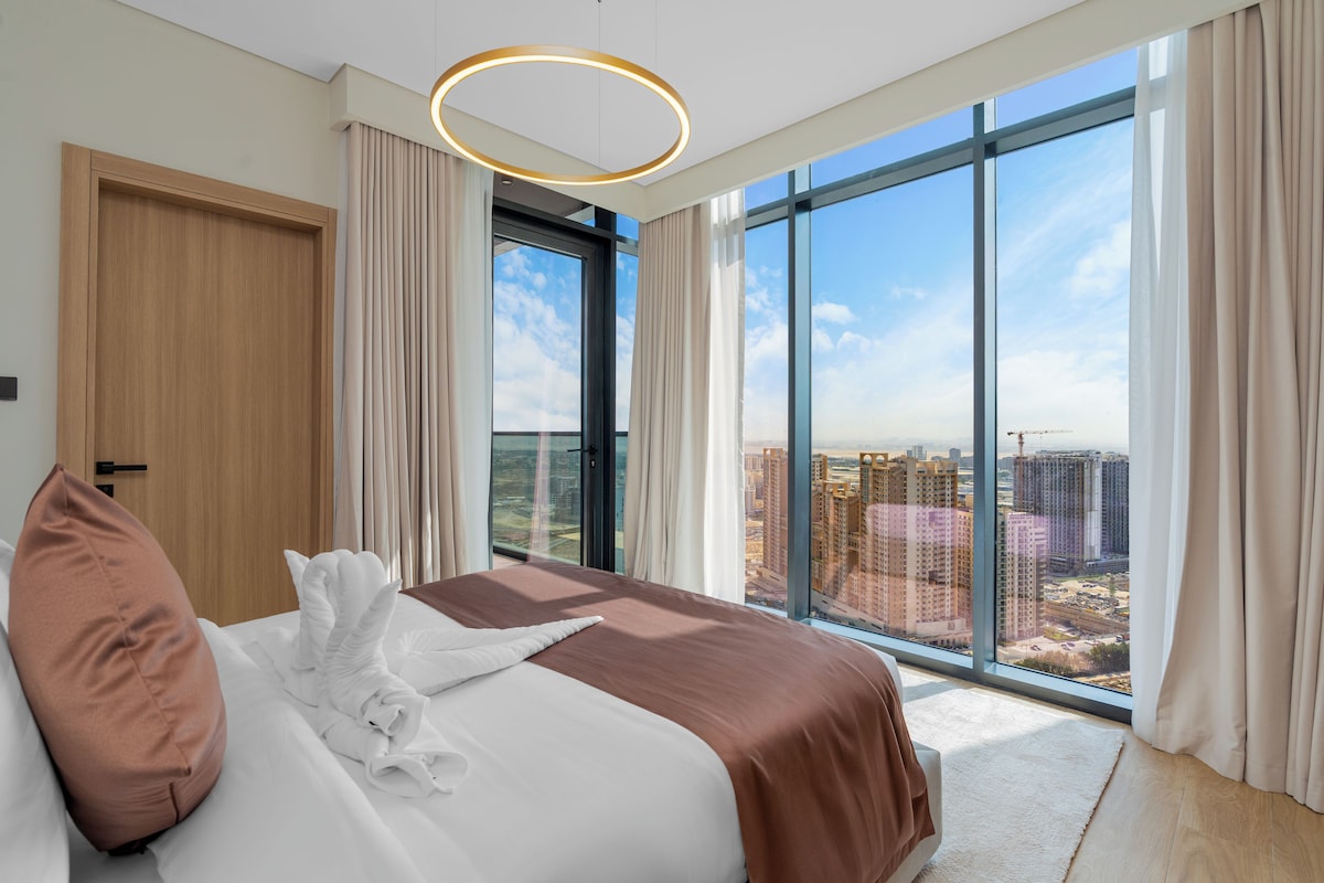 Quarto de hotel com uma cama grande, porta de madeira e vista da cidade através de janelas do chão ao teto.