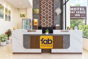 Bancada de recepção de hotel com placa 'fab hotels', placa de 'proibido fumar' e área de recepção com divisória decorativa e obras de arte.