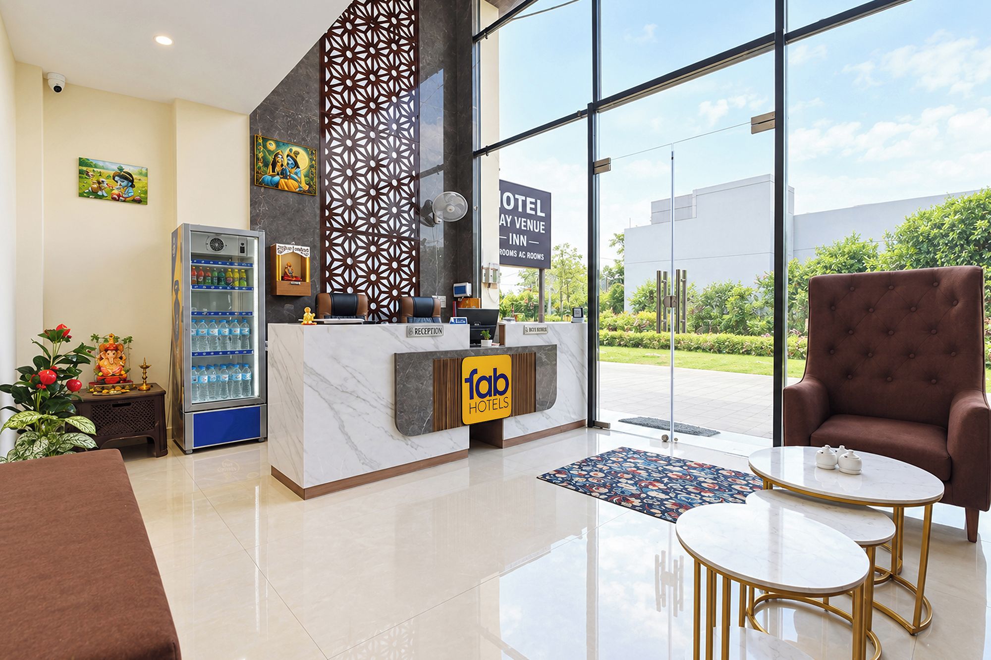 Um saguão de hotel com balcão de mármore, mesa de atendimento, área de estar com poltrona marrom e uma pequena mesa, e uma parede decorativa com arte religiosa.