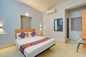 Quarto de hotel com cama, mesa de cabeceira, luminária, aparelho de ar condicionado e uma porta que dá para outro cômodo.