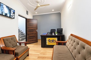 Um saguão de hotel com um balcão de recepção, dois sofás, um ventilador de teto e uma TV fixada na parede.