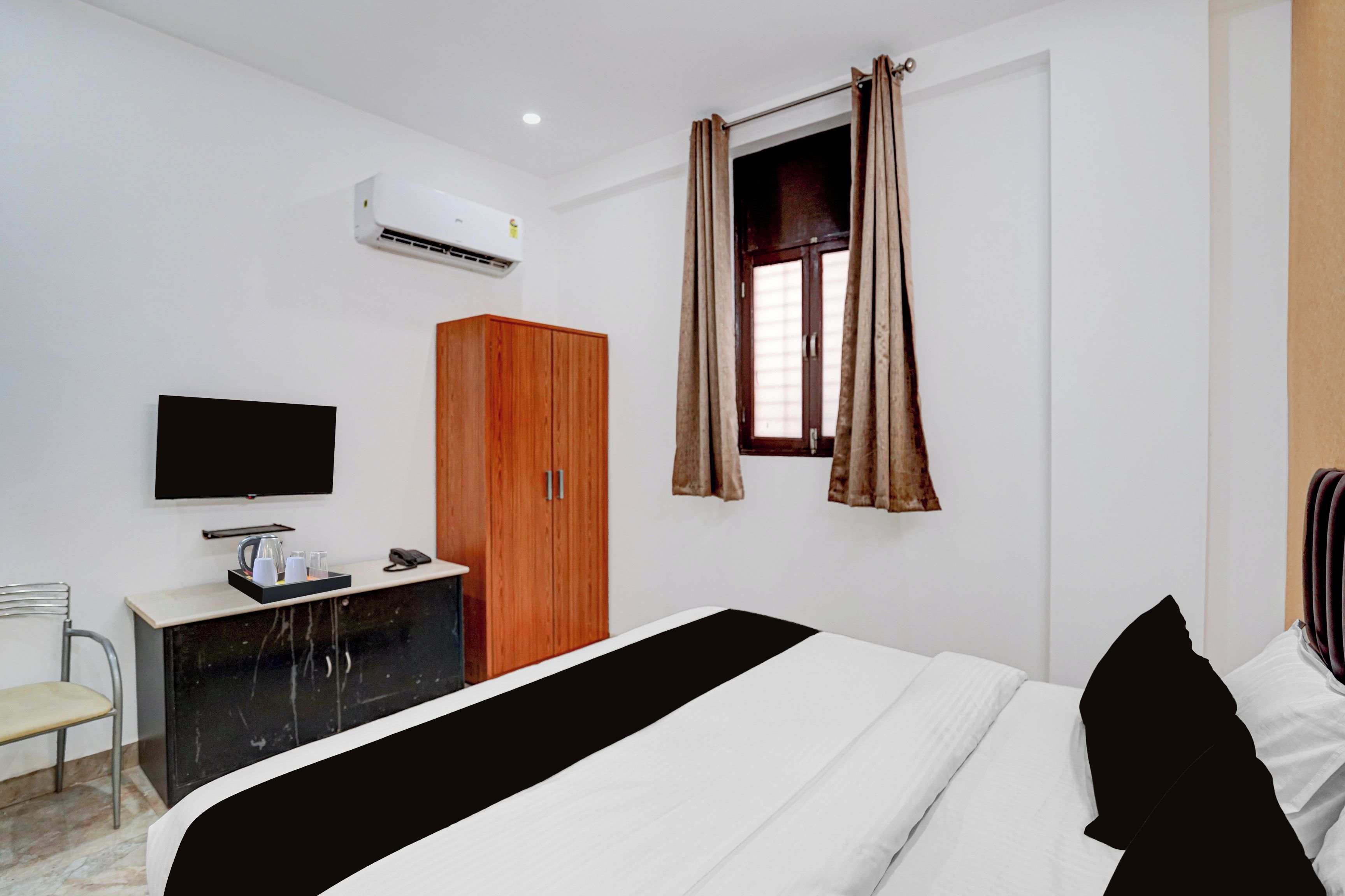 Quarto de hotel com cama, televisão, guarda-roupa, secretária com telefone e máquina de café, e janela com cortinas.