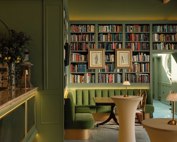 Uma biblioteca com estantes, um sofá verde, uma mesa e duas molduras.