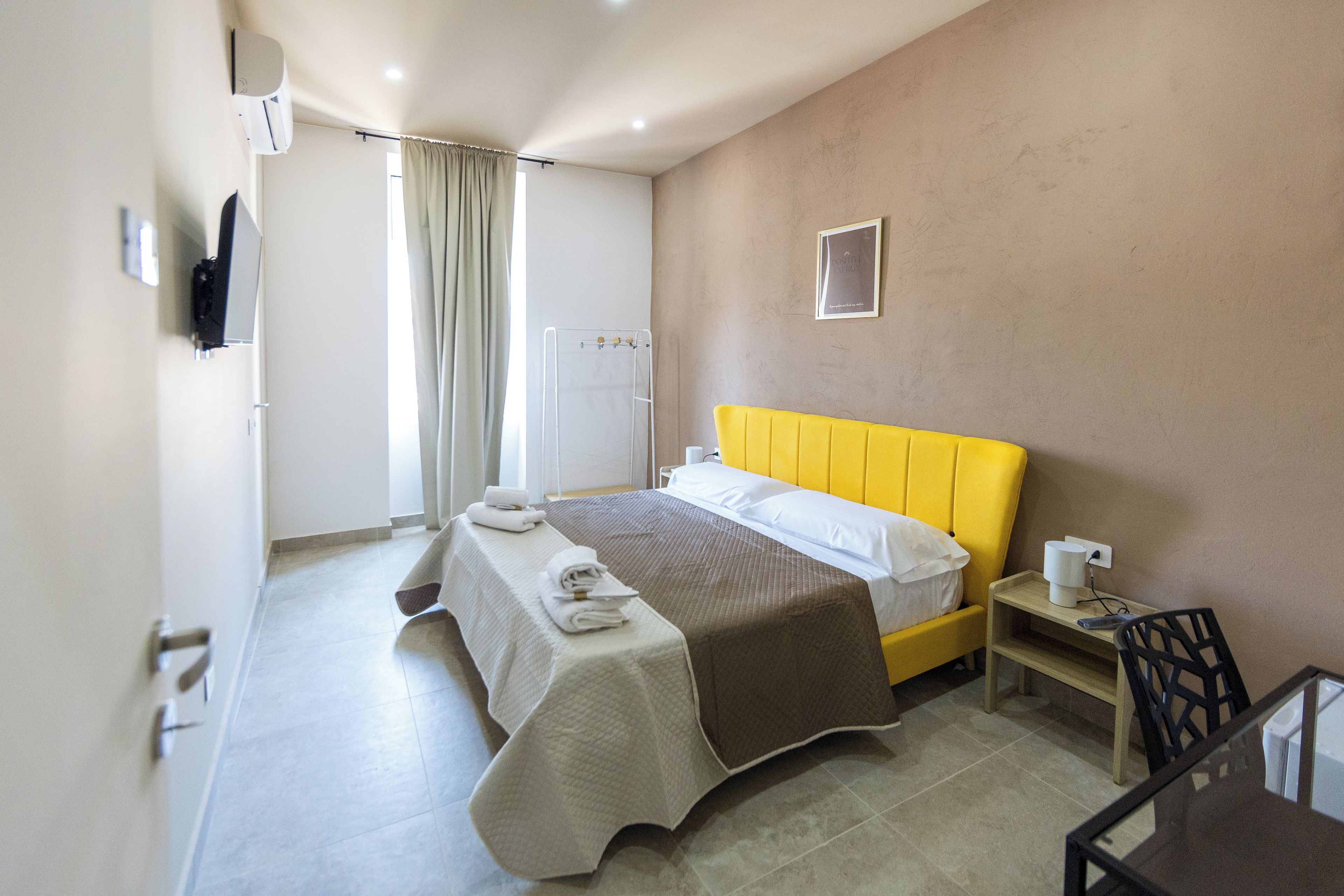 Quarto de hotel com cama, mesa de cabeceira, cadeira, janela com cortinas e ar condicionado fixado na parede.