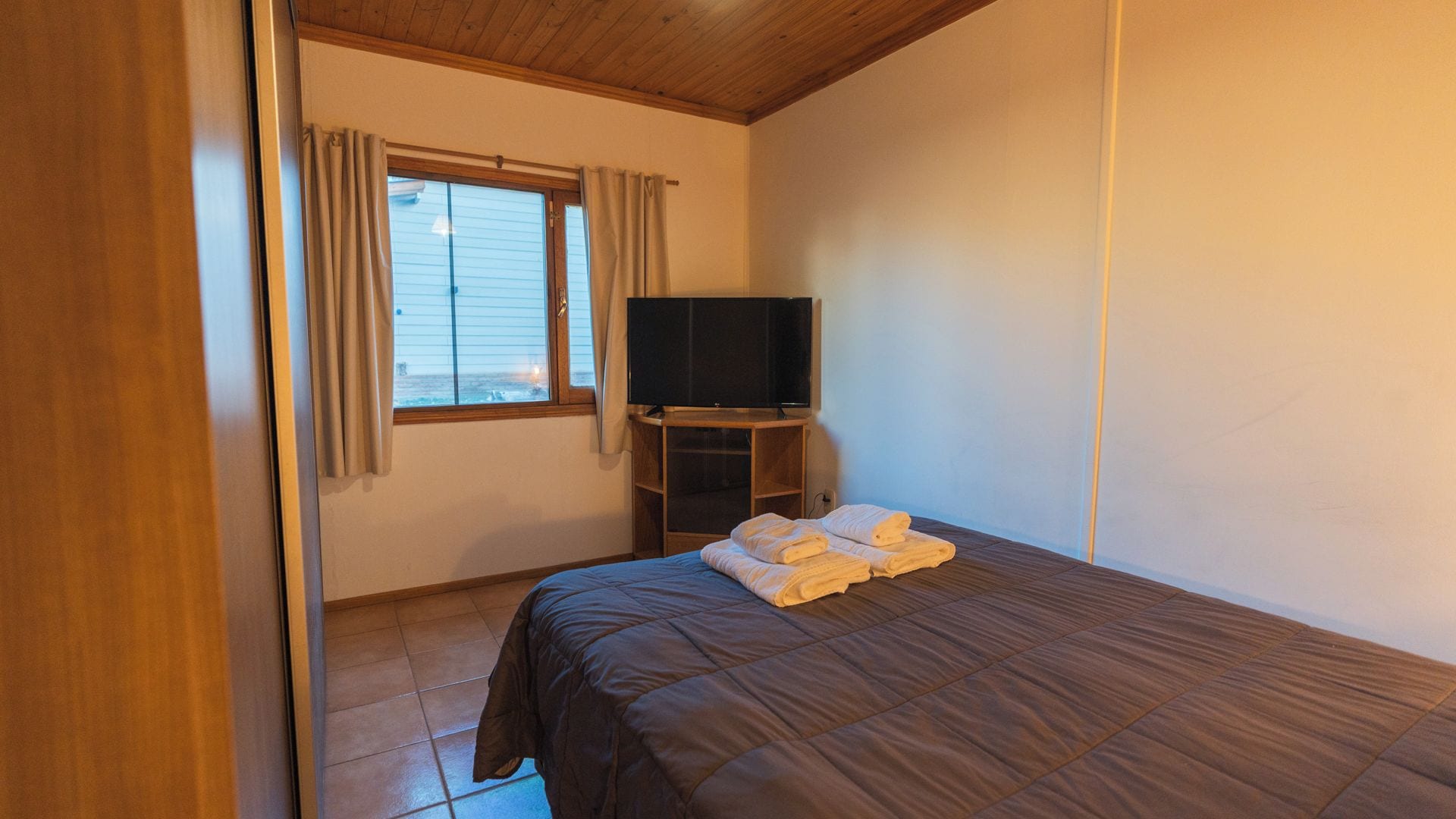 Quarto com cama, televisão em suporte de madeira e janela com cortinas.