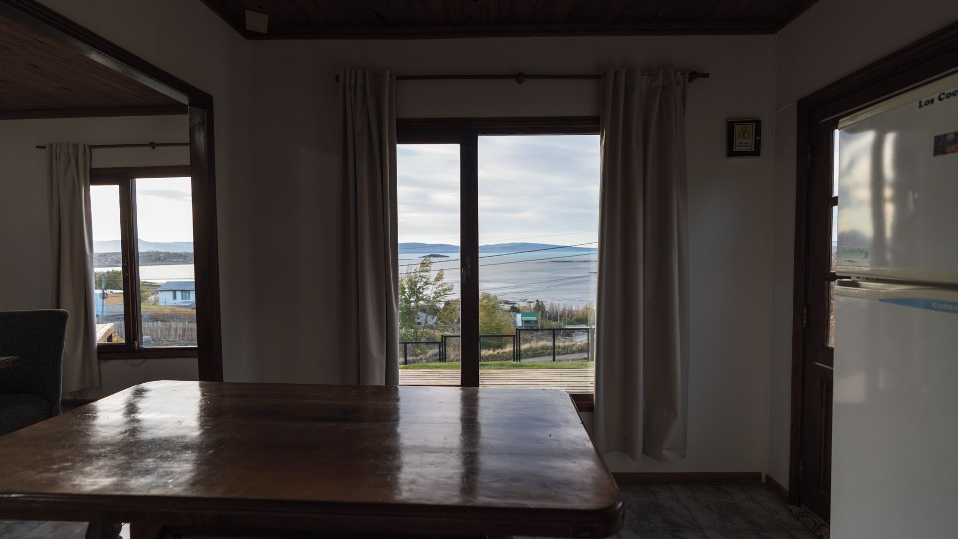 Quarto com mesa de madeira, vista para um corpo d'água e porta que dá para o exterior.
