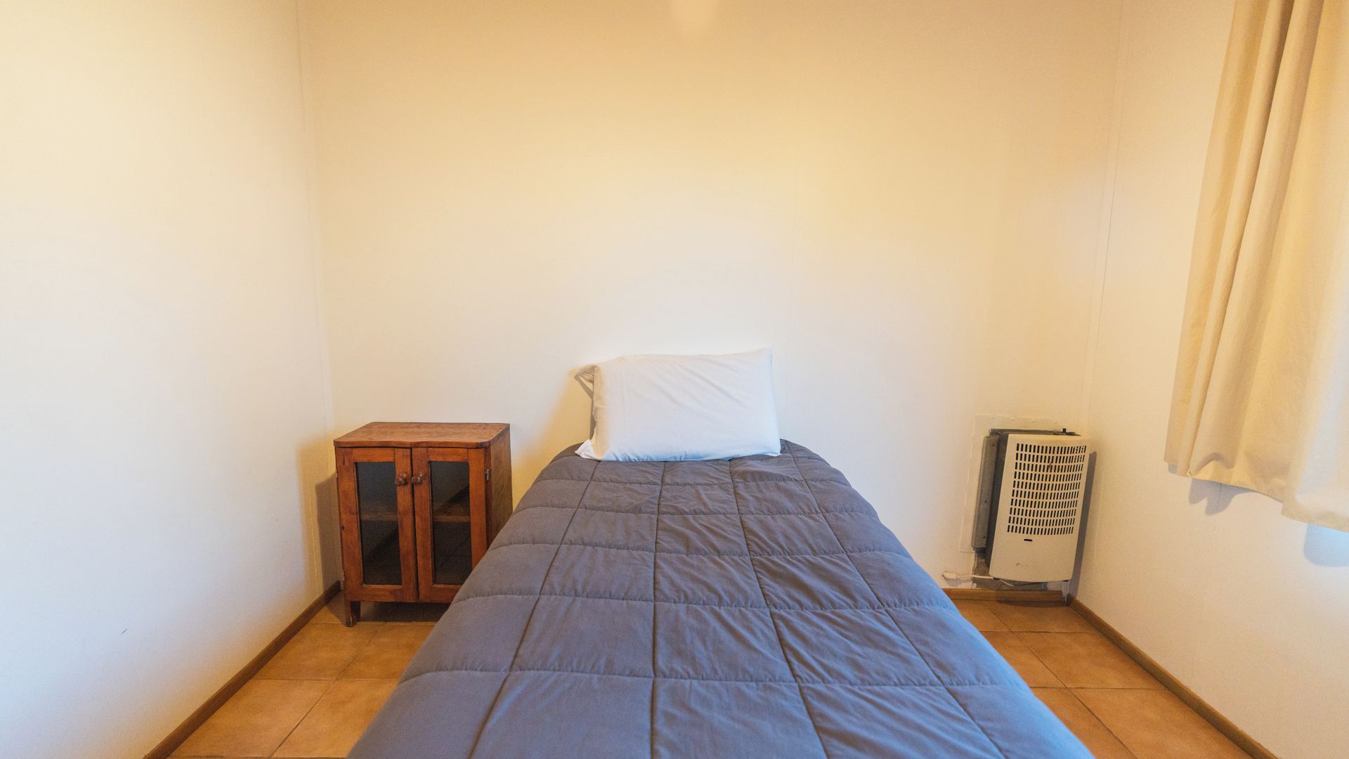 Quarto pequeno e estreito com uma cama, um armário de madeira e um aquecedor.