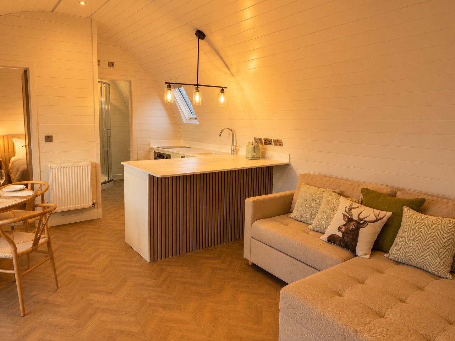 Um interior acolhedor com piso de madeira, um sofá com almofadas estampadas, uma área de jantar com mesa e cadeiras, e uma cozinha com ilha central.