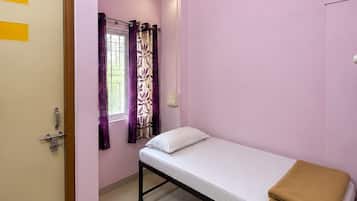 Quarto individual com cama, ventoinha no teto, janela com cortinas roxas e uma pequena mesa com uma planta.