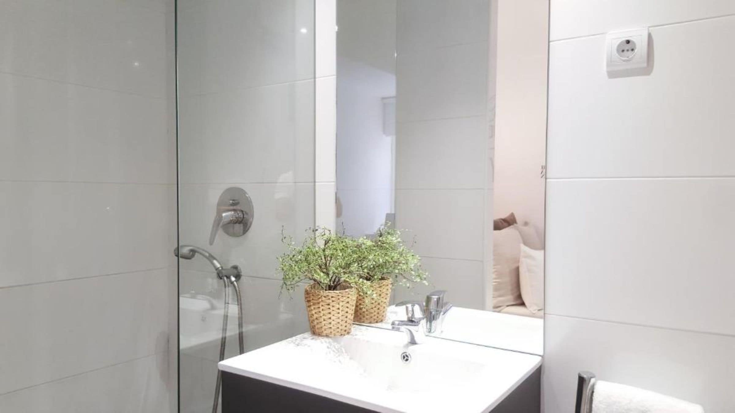 Casa de banho moderna com chuveiro em cabine de vidro, lavatório branco e duas plantas em vasos.