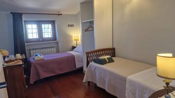 Quarto com duas camas, um radiador, janela com cortinas, uma mesa com abajur e um guarda-roupa.