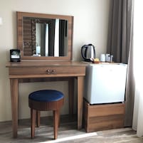 Quarto compacto com escrivaninha de madeira, um banquinho, frigorífico pequeno, chaleira e espelho.