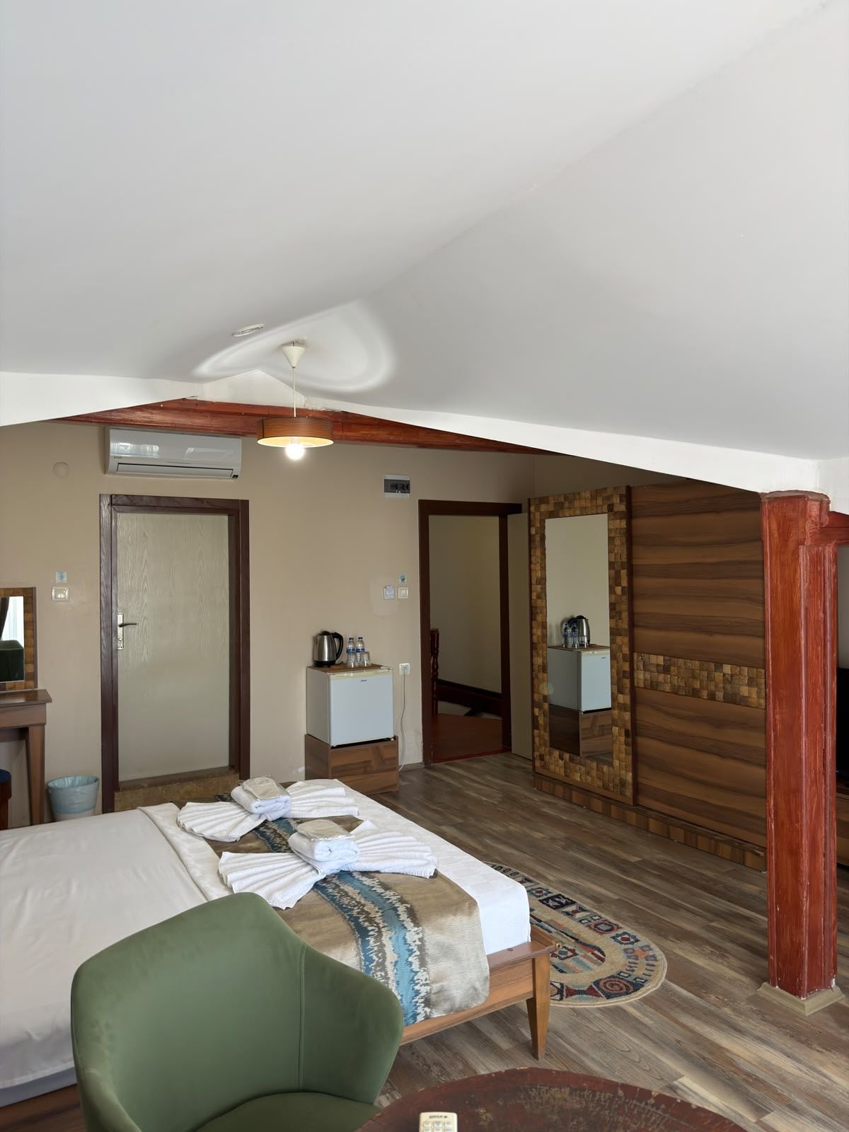 Quarto de hotel com cama, uma cadeira verde, mesa pequena, espelho e painel de parede de madeira.
