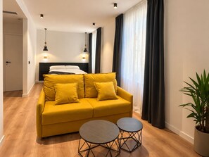 Quarto de hotel moderno com sofá amarelo, duas mesas redondas, cama com cabeceira escura e planta em vaso.