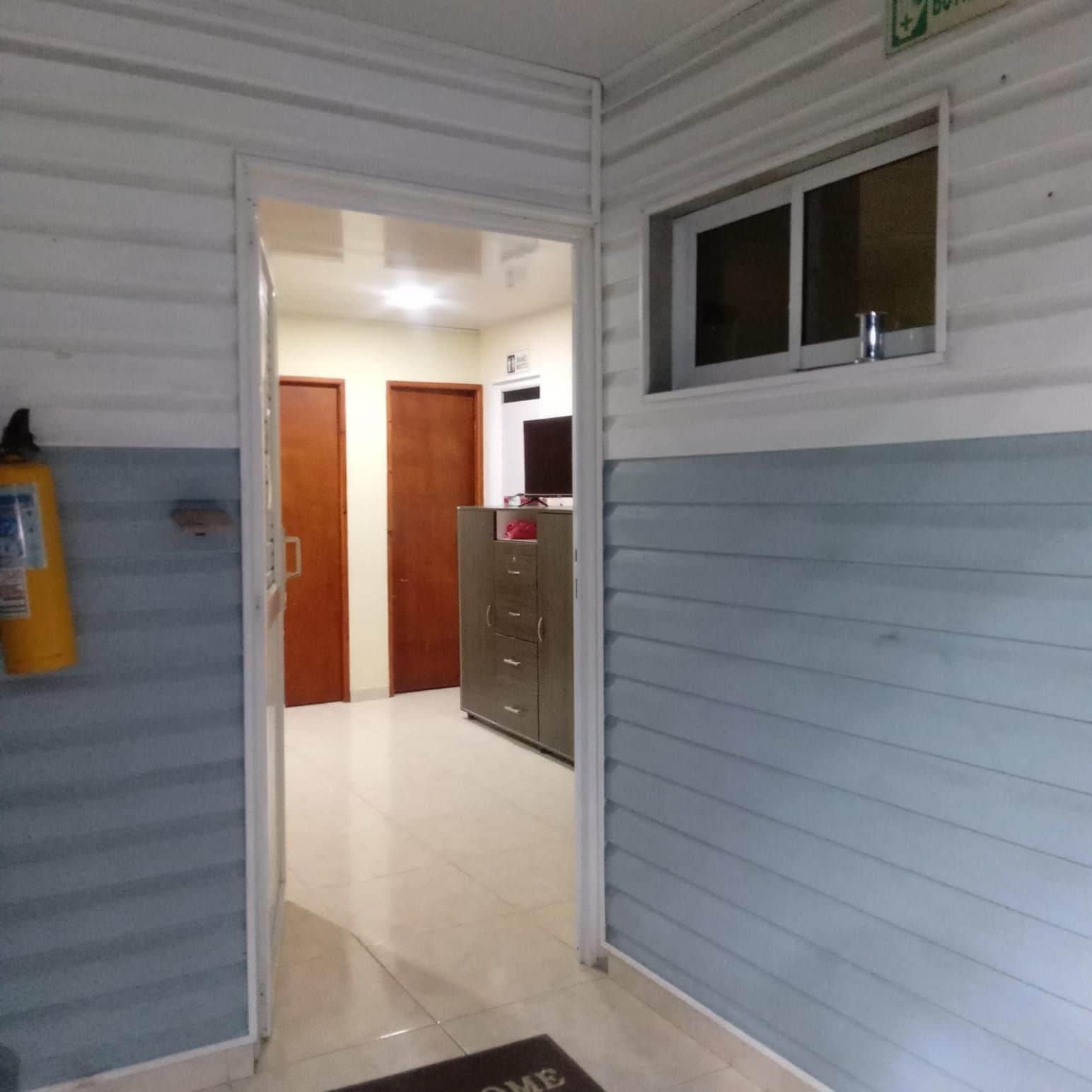 Um corredor com uma porta que dá para um quarto, um extintor de incêndio e uma janela com vista.