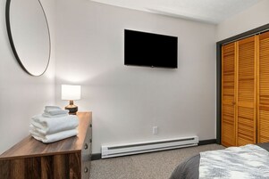Quarto de hotel com televisão de tela plana na parede, mesa de cabeceira com abajur e toalhas, aquecedor sob a janela e armário de madeira.