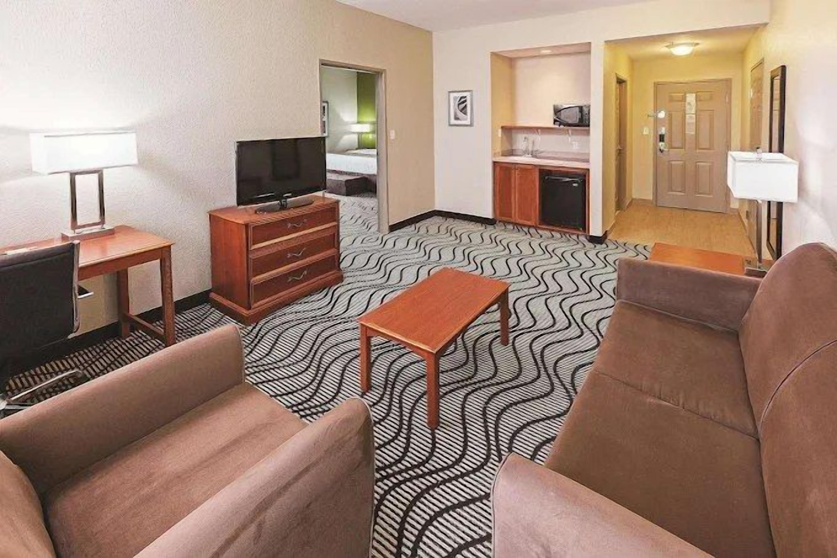 Quarto de hotel com sofá, televisão, mesa pequena, cozinha compacta e tapete estampado.