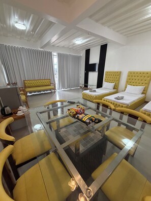 Quarto de hotel moderno com mesa de jantar de vidro, cadeiras estofadas amarelas, um sofá e duas camas com cabeceiras amarelas.