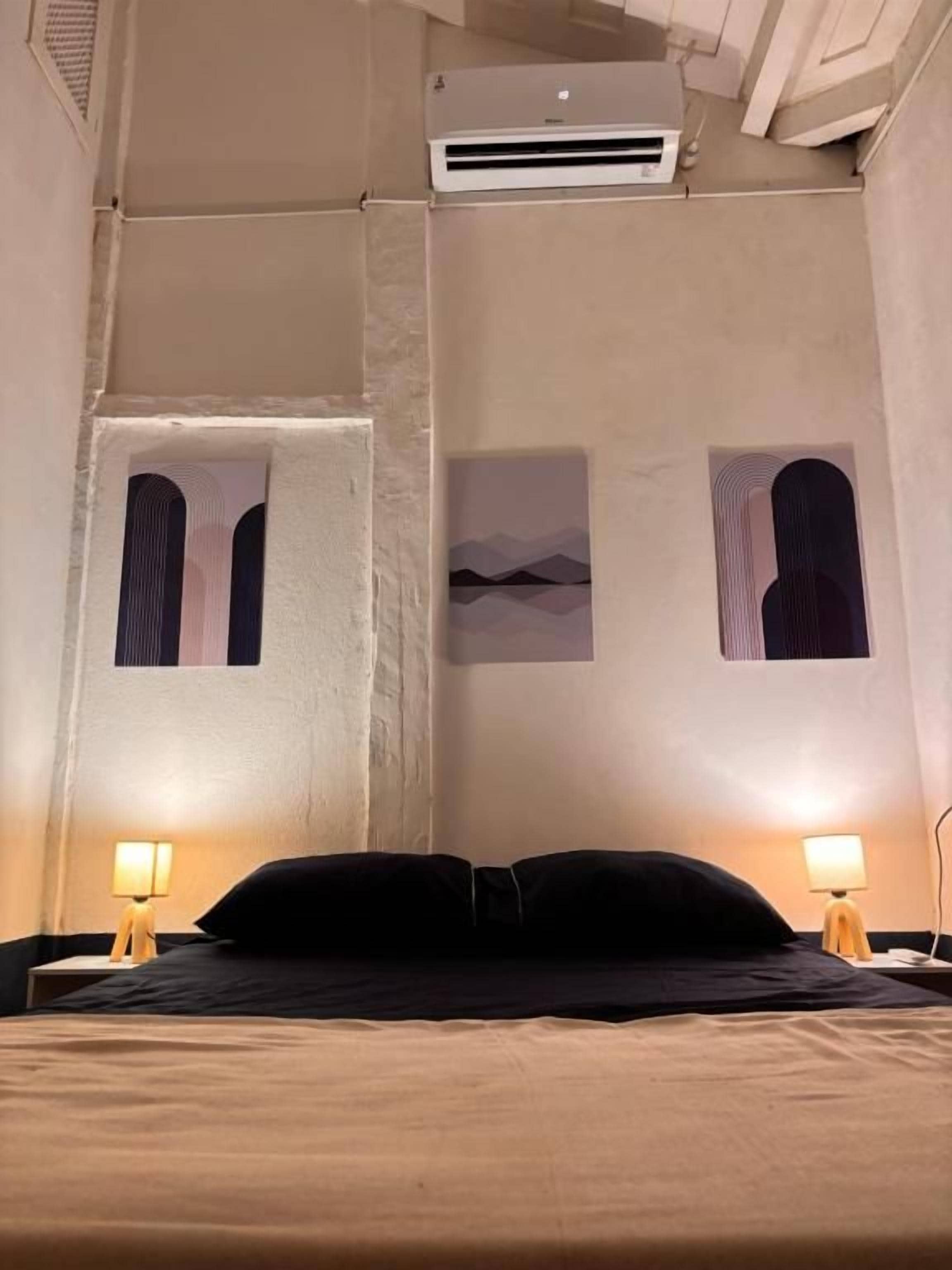 Quarto de hotel com uma cama, duas luminárias de cabeceira, um aparelho de ar condicionado e três quadros pendurados na parede.