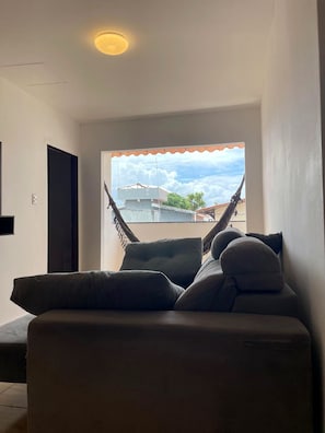 Quarto com rede, sofá e vista para prédios pela janela.