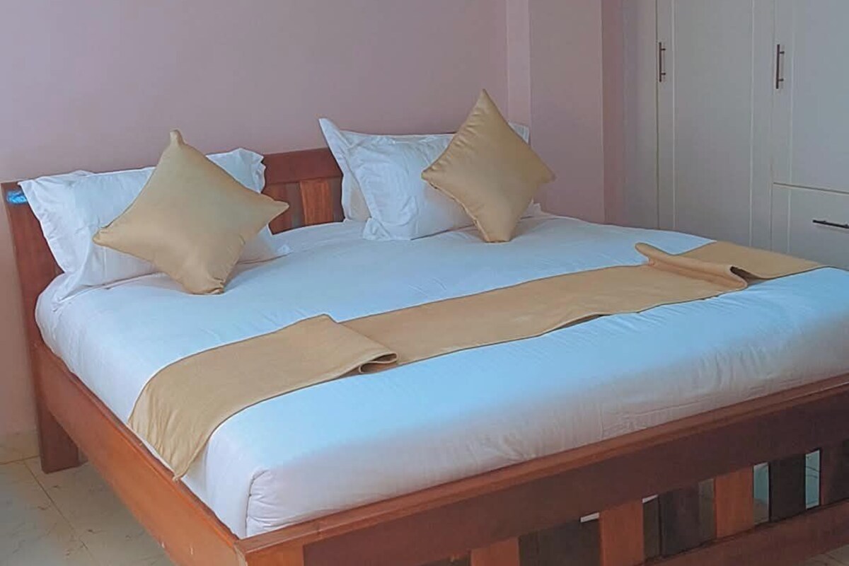 Uma cama com estrutura de madeira, roupa de cama branca e almofadas bege em um quarto com paredes rosa e porta branca.
