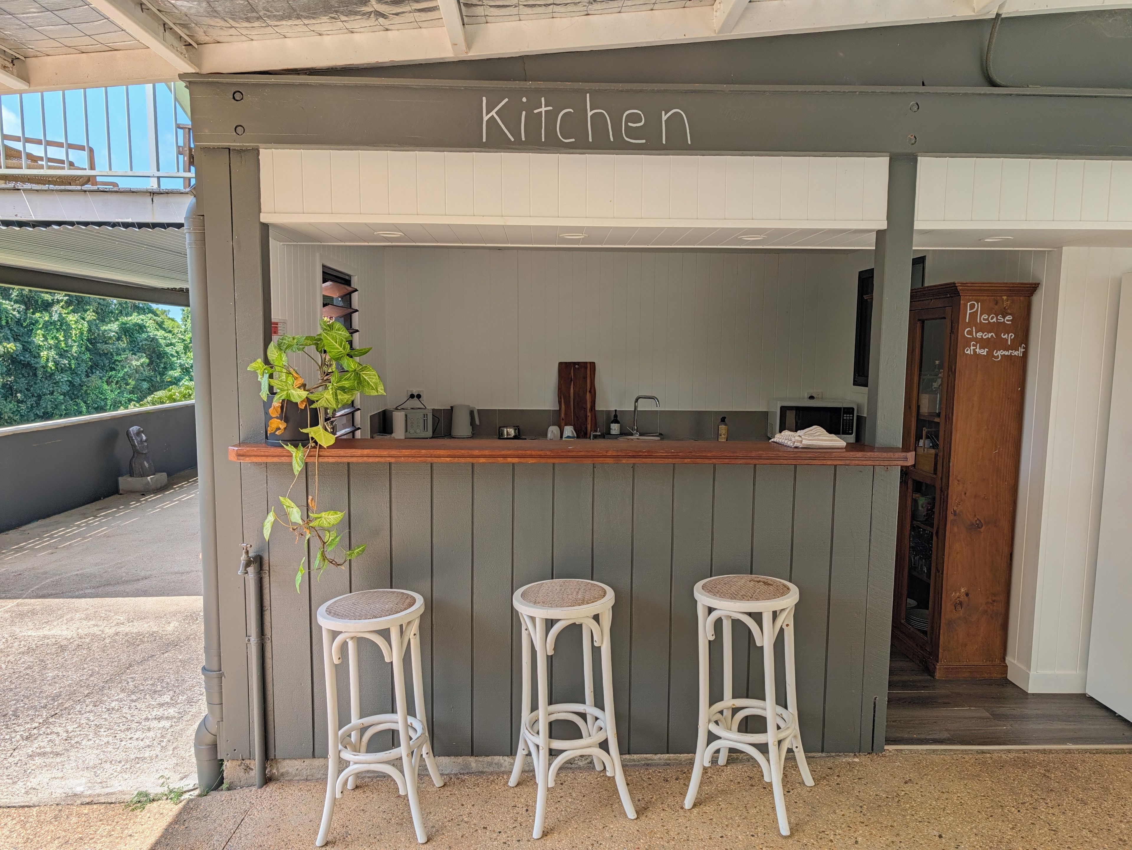 Uma área de cozinha com uma placa indicando 'Cozinha', três banquetas brancas, um balcão de madeira e uma placa solicitando aos hóspedes que limpem após o uso.