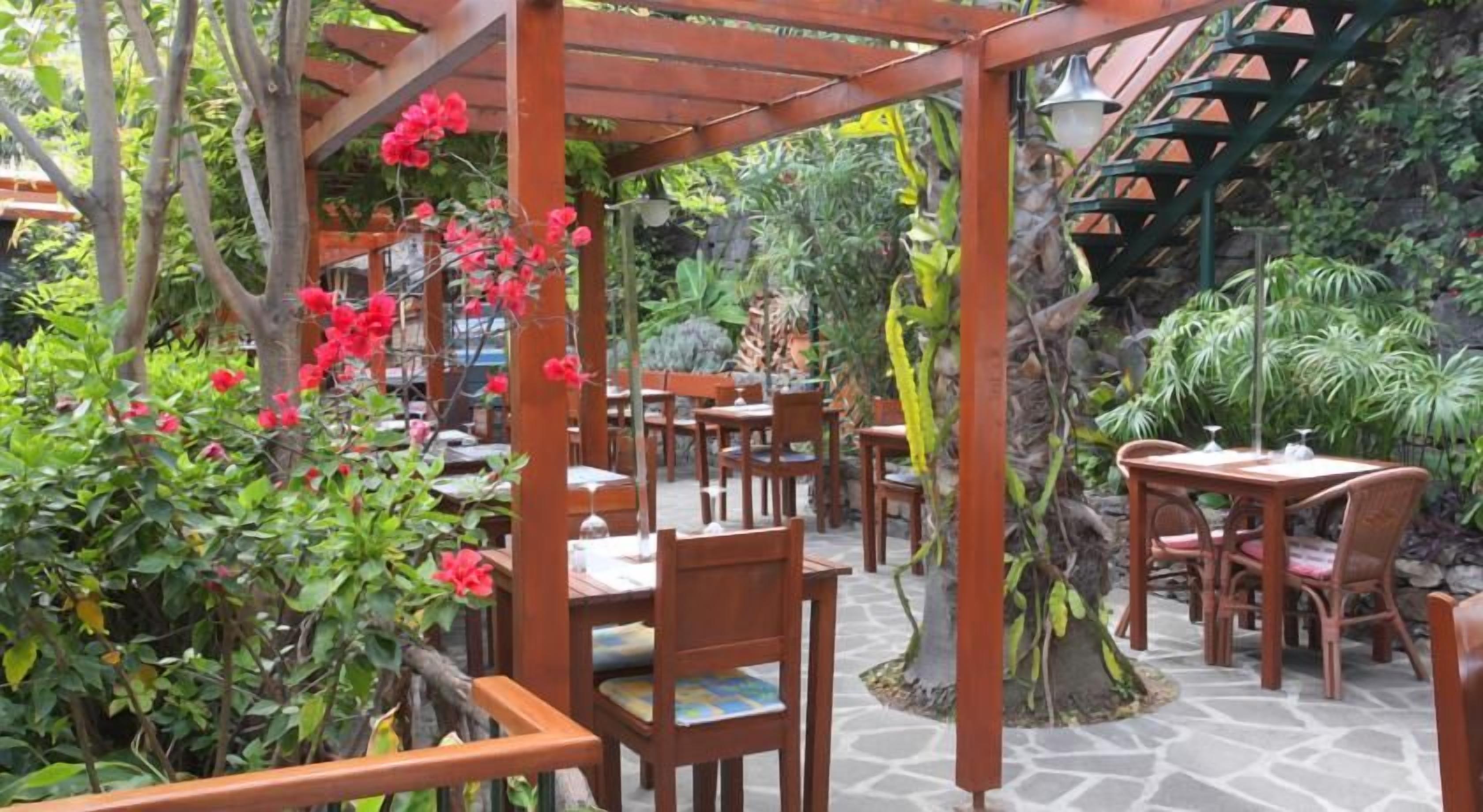 Pátio externo com mesas e cadeiras de madeira, rodeado por vegetação exuberante e plantas floridas.