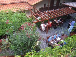 Uma área de pátio externa com pérgulas de madeira, mesas e cadeiras, cercada por folhagem exuberante e plantas em vasos.